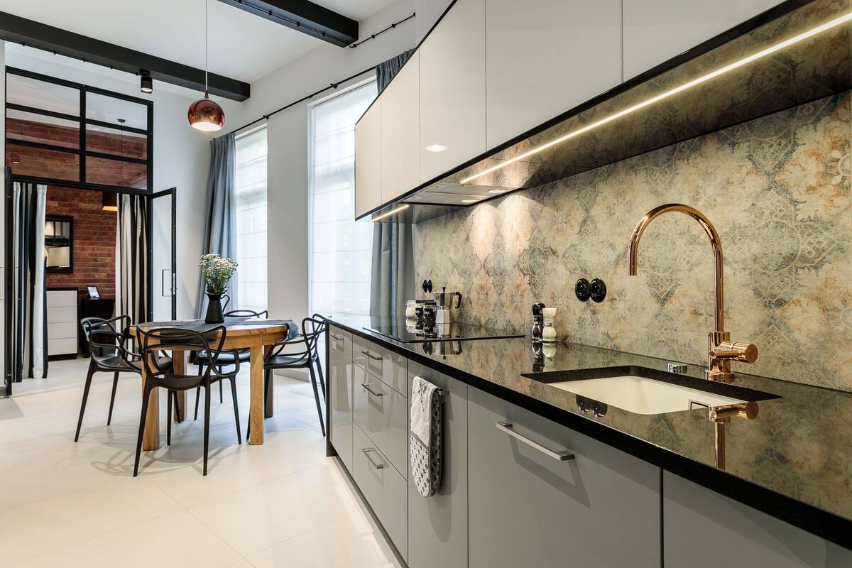 Cozinha moderna com painel de parede em mármore, torneira dourada e área de jantar com mesa e cadeiras de madeira.