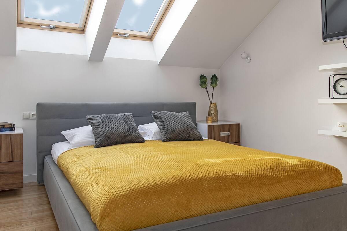 Quarto moderno com claraboia, uma cama com manta cor mostarda e uma mesa de cabeceira com um vaso e livros.