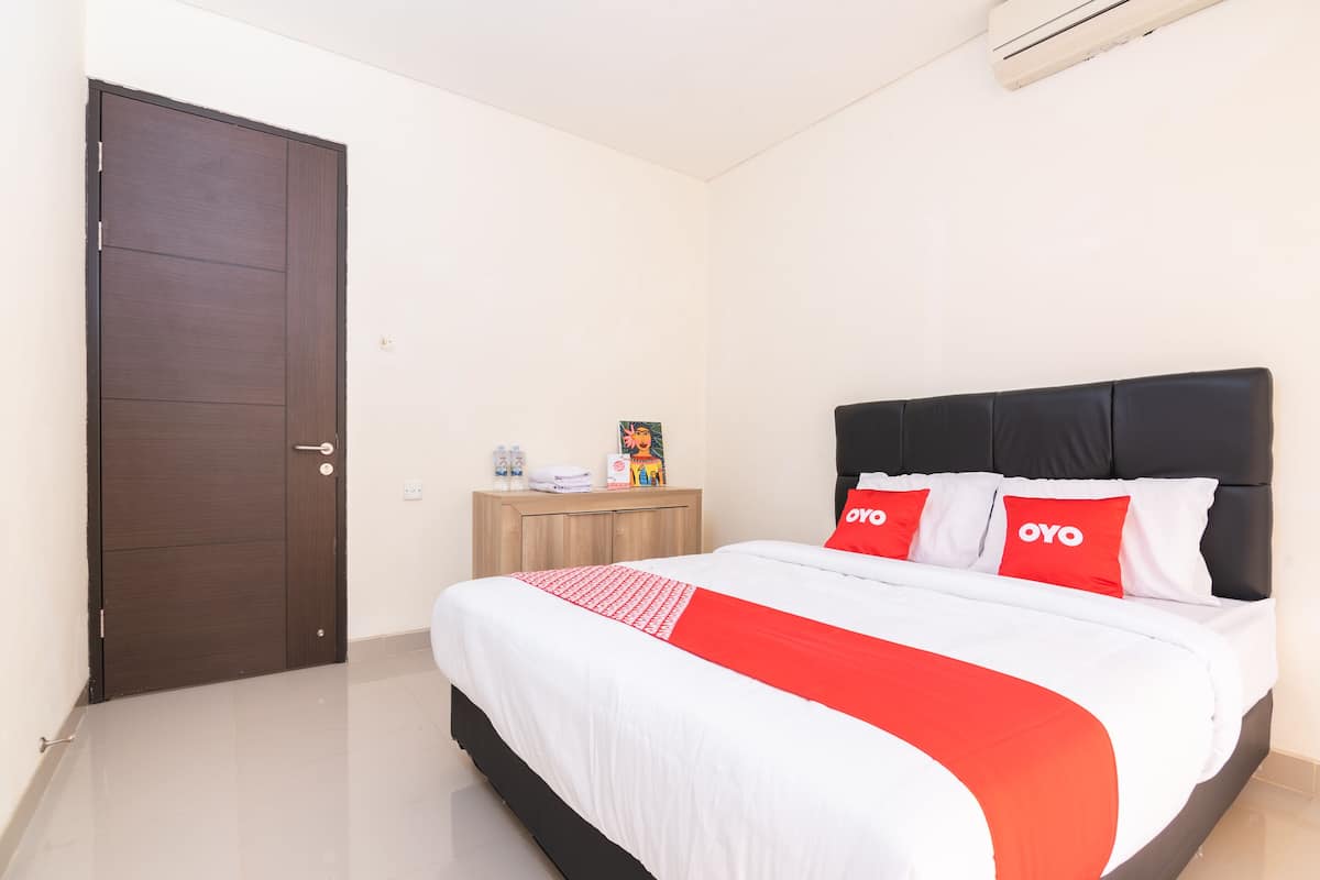 Quarto de hotel com uma cama, uma mesinha de cabeceira de madeira, uma parede branca e uma porta castanha.