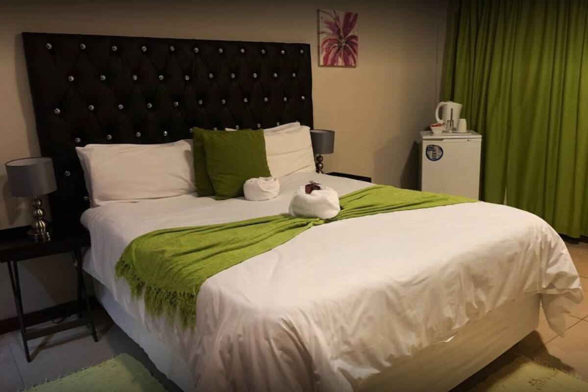 Uma cama bem arrumada com lençóis brancos e um cobertor verde, uma mesinha de cabeceira com abajur, e um pequeno armário branco com cafeteira.