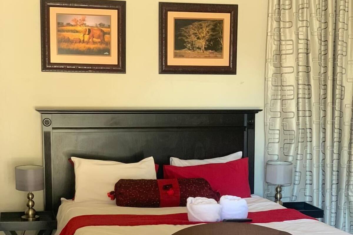 Uma cama bem arrumada com roupa de cama vermelha e branca, uma poltrona marrom e duas molduras na parede.