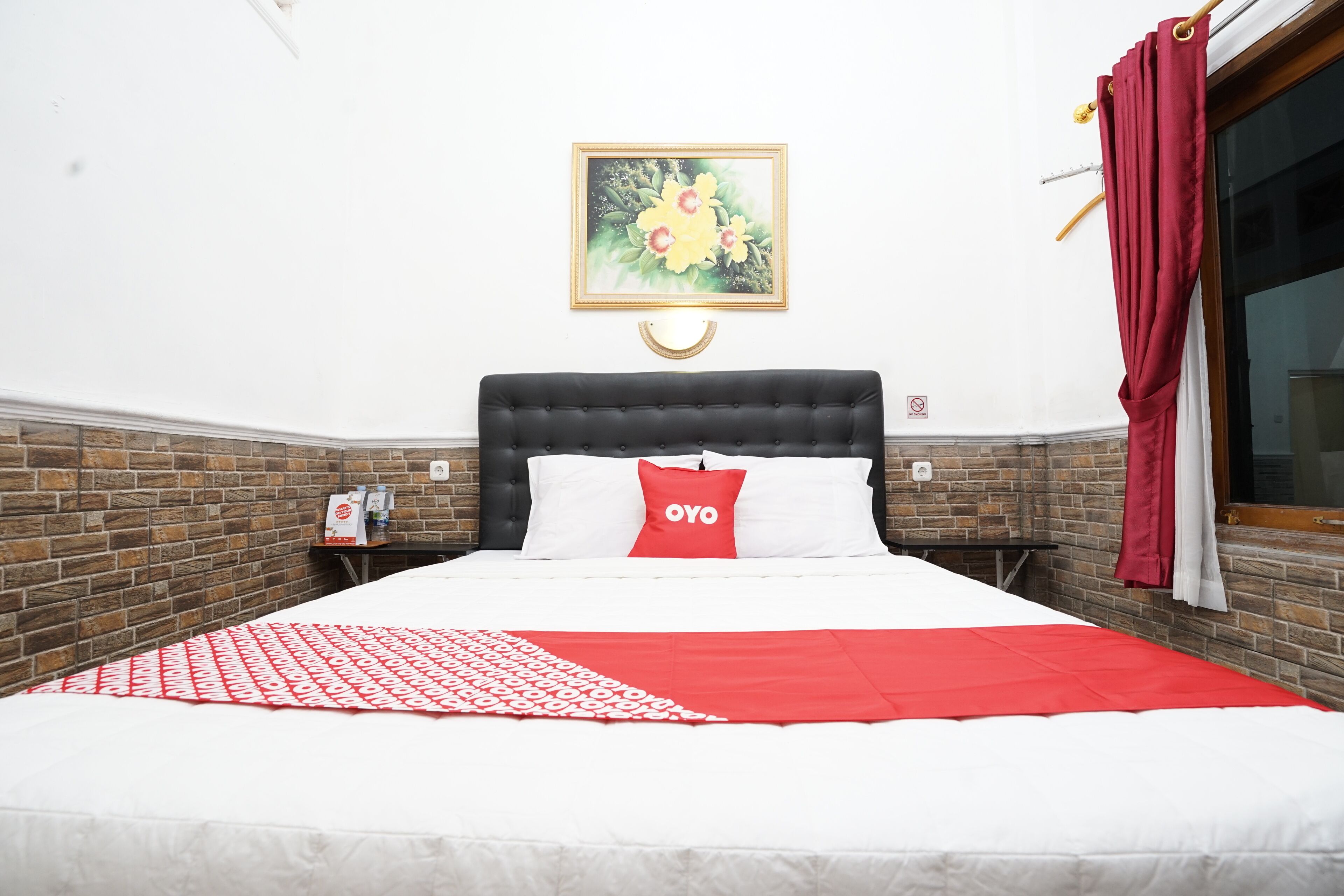 Uma cama bem arrumada com um cobertor estampado em vermelho e branco, uma cabeceira preta e um único travesseiro vermelho com a palavra "OYO" escrita nele.