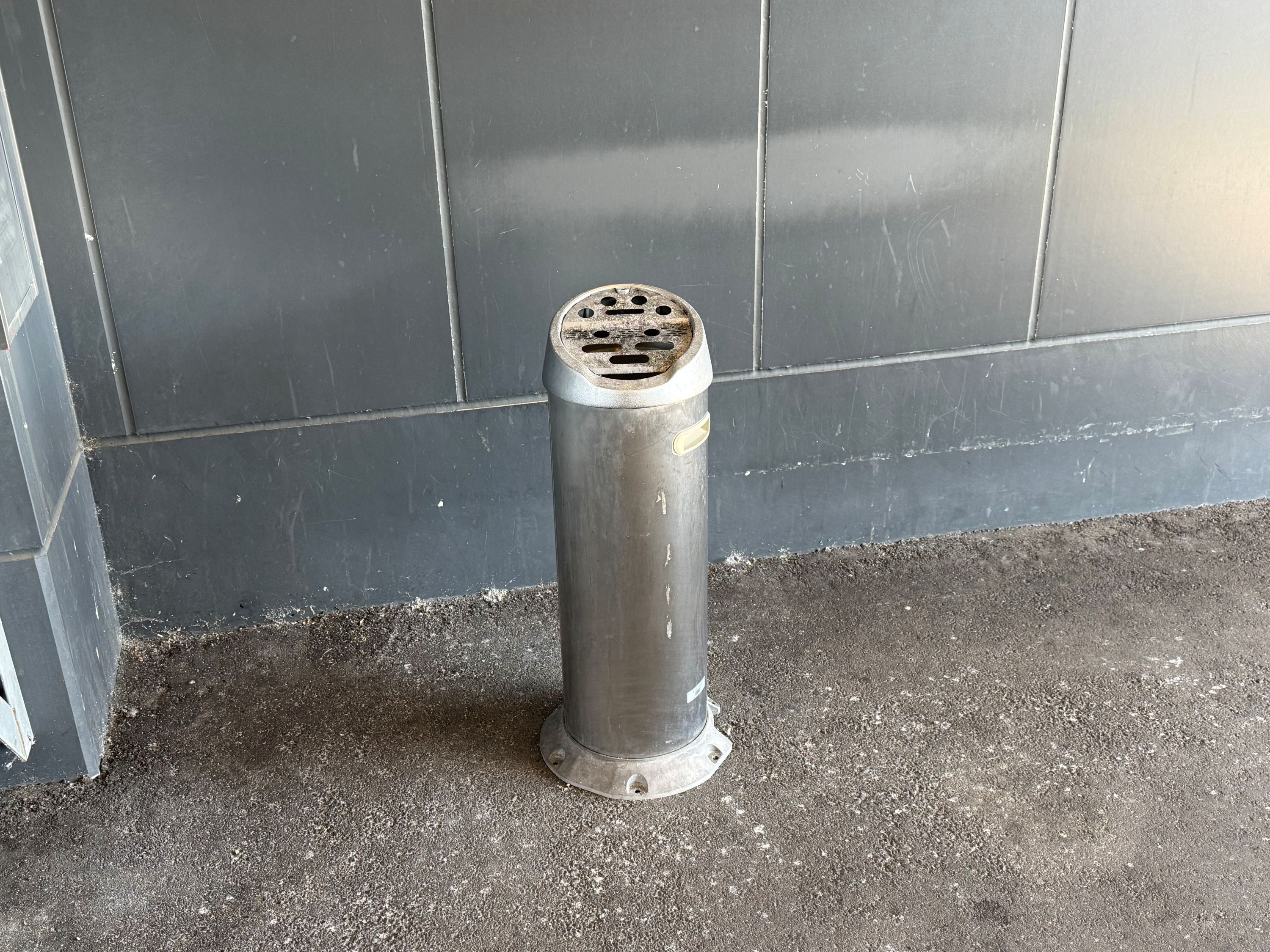 Um poste de metal com topo circular e furos, fixado sobre superfície de concreto em frente a uma parede cinzenta.