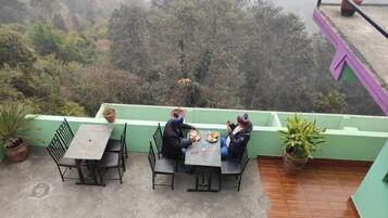 Terraço com mesa e cadeiras, rodeado por plantas em vasos e vista para uma área arborizada.