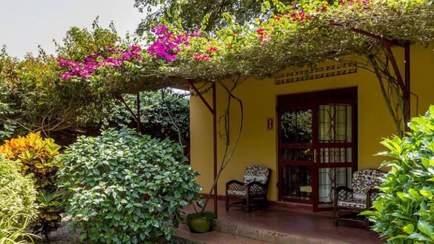 Um edifício amarelo com varanda coberta, duas cadeiras e um vaso de planta.