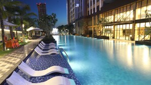 Piscina com espreguiçadeiras e guarda-sóis, cercada por um edifício com grandes janelas e uma palmeira.