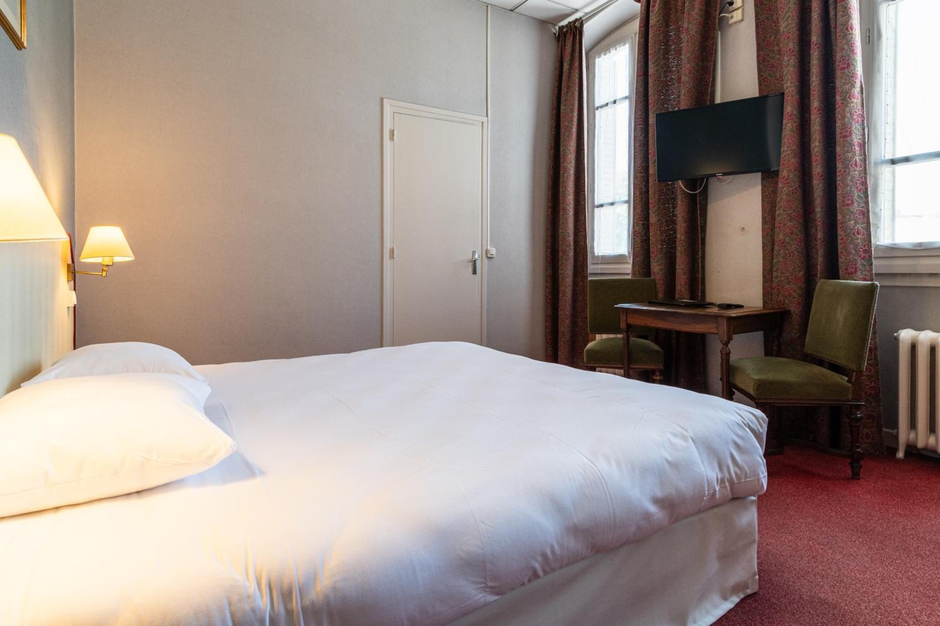Quarto de hotel com uma cama grande, uma mesa com cadeira, televisão e janela com cortinas.