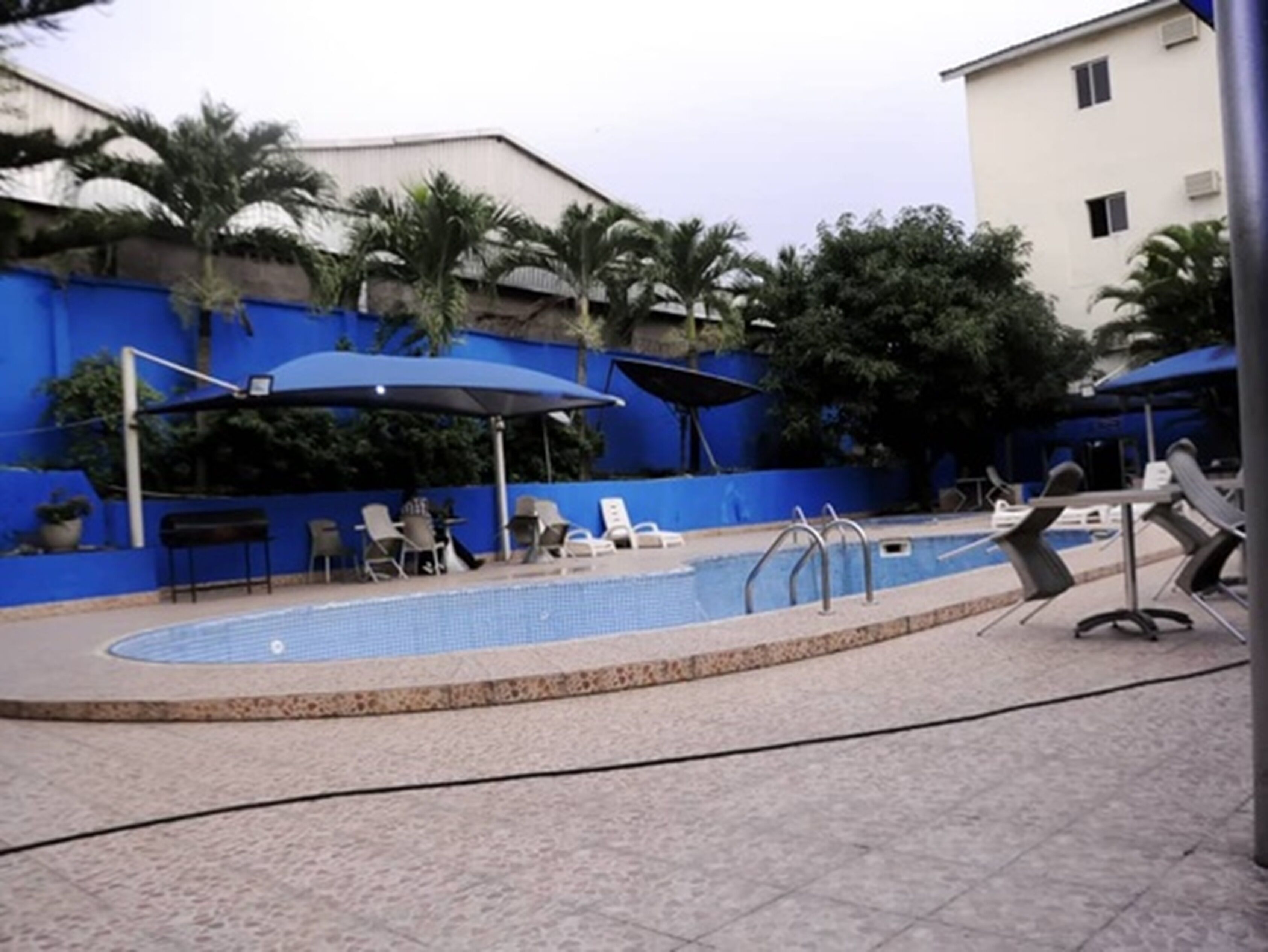 Piscina externa com espreguiçadeiras e guarda-sóis, cercada por um muro azul e palmeiras.