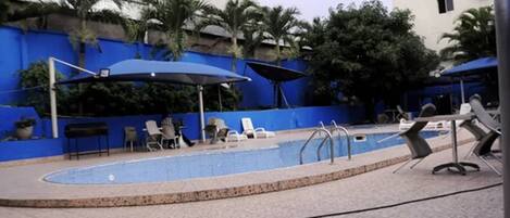 Piscina externa com espreguiçadeiras e guarda-sóis, cercada por um muro azul e palmeiras.