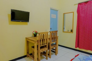 Quarto com escrivaninha de madeira, duas cadeiras, televisão na parede, porta, espelho e cortina vermelha.