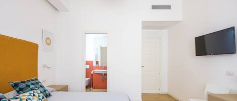 Quarto moderno com cama, televisão, escrivaninha e banheiro visível através de uma porta aberta.