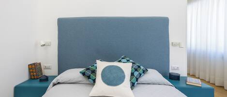 Cama bem arrumada com cabeceira azul, duas mesas de cabeceira com livros e uma pequena planta, e uma cortina branca na janela.