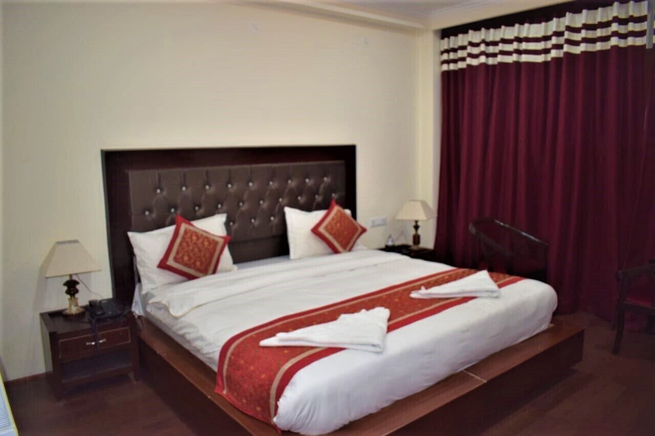 Quarto de hotel com uma cama grande, uma mesa de cabeceira, uma luminária e uma janela com cortinas.