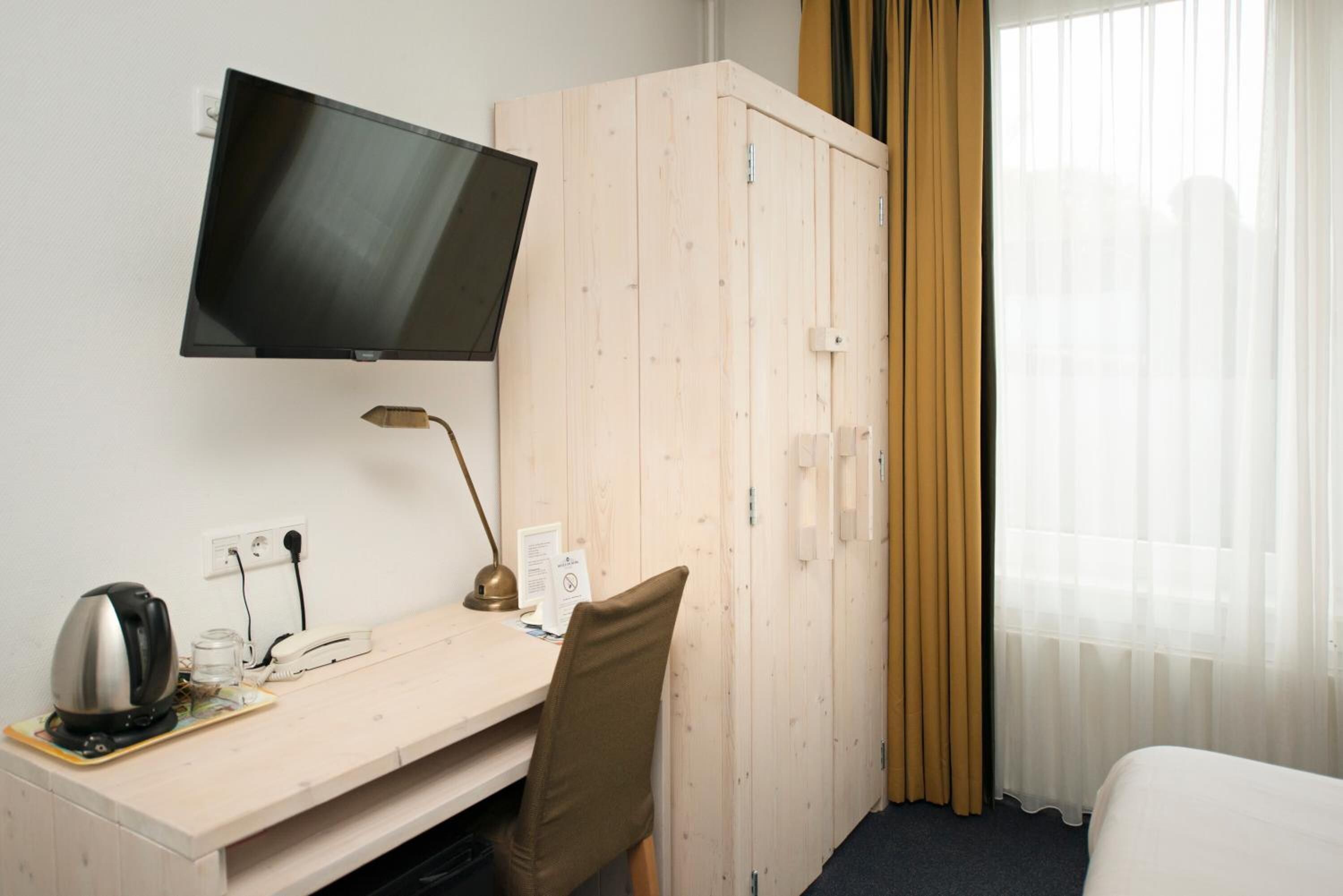 Quarto de hotel com uma escrivaninha de madeira, televisão, cadeira, guarda-roupa e janela com cortinas.