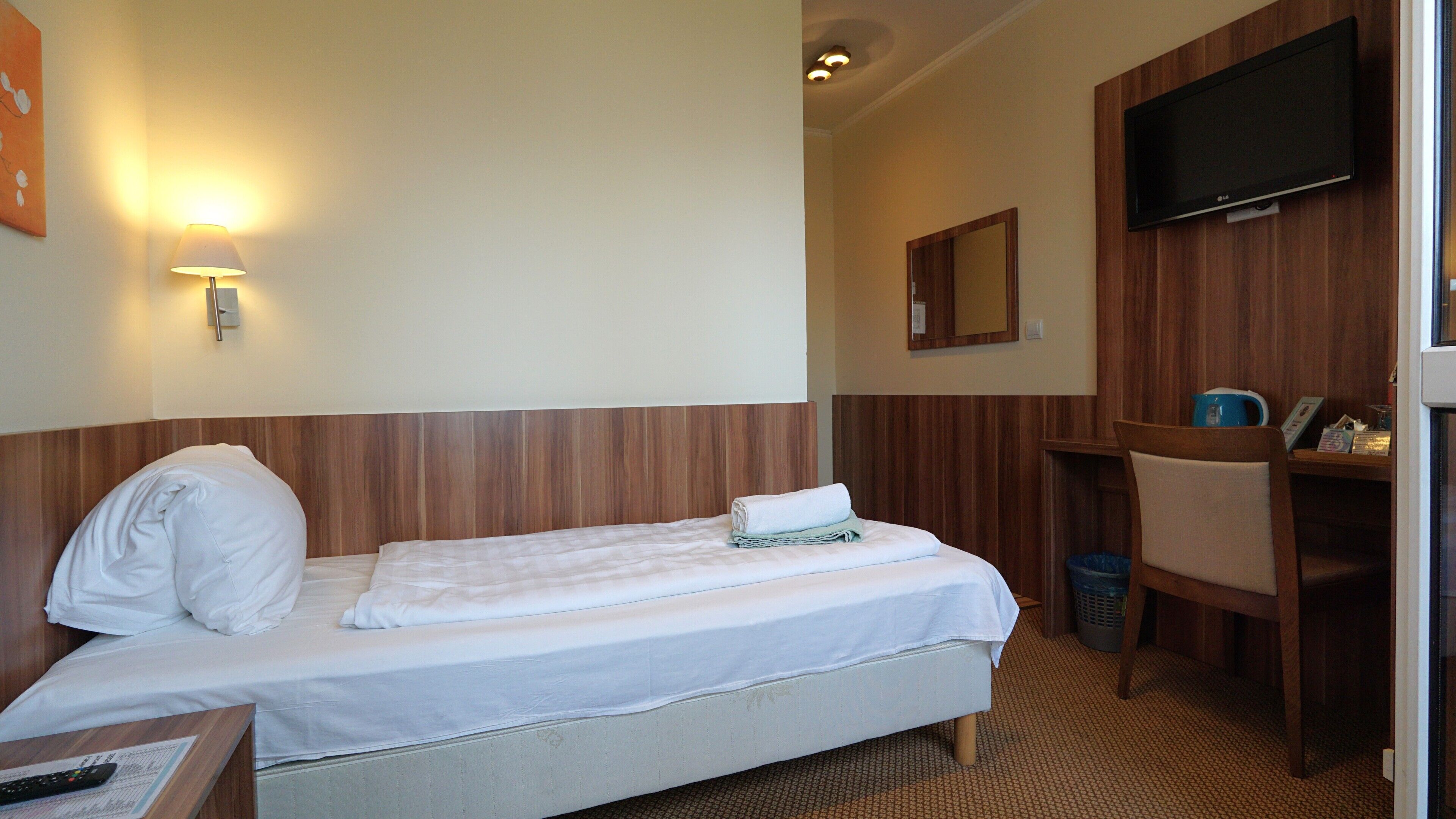 Quarto de hotel com cama, escrivaninha, cadeira, televisão e espelho fixado na parede.