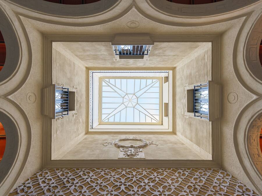 Um amplo átrio com claraboia central, colunas ricamente decoradas e piso padronizado.
