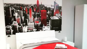 Quarto de hotel com um grande mural da paisagem urbana na parede, uma cama com manta vermelha e uma mesa de cabeceira branca.