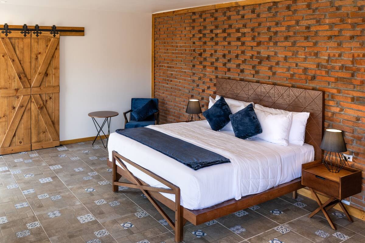 Quarto com cama de madeira, parede de tijolos, uma mesa pequena e uma cadeira.