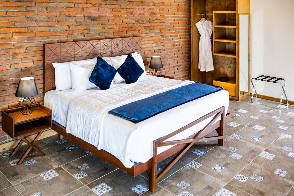 Cama bem arrumada com estrutura de madeira, duas mesas de cabeceira com abajures, manta azul e parede de tijolos ao fundo.