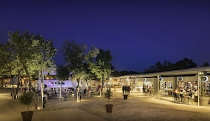 Um espaço de café ao ar livre, com mesas e cadeiras, cercado por árvores e iluminado com iluminação ambiente.