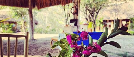 Um ambiente rústico ao ar livre com uma estrutura de telhado de palha, uma mesa com um vaso de flores e uma cadeira.