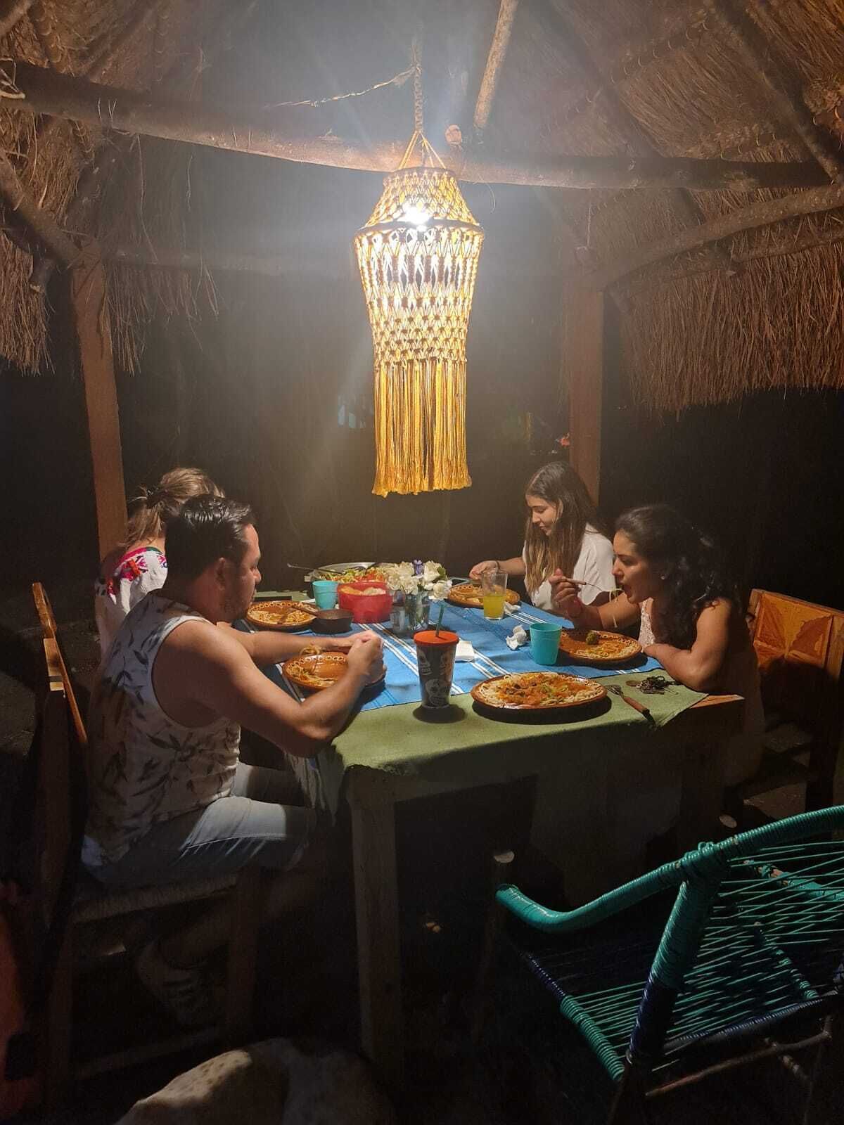 Um grupo de pessoas jantando à mesa sob um telhado de palha, com um candeeiro pendurado e uma cadeira de fibra natural.
