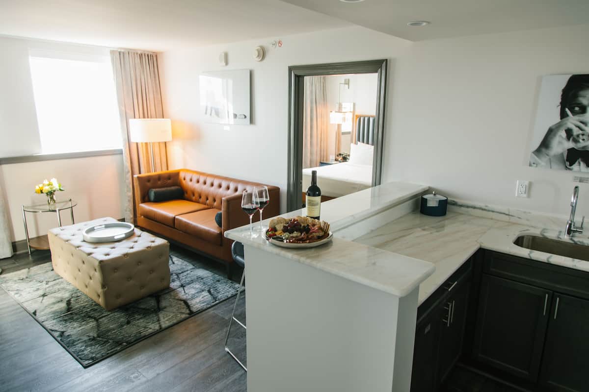 Quarto de hotel moderno com área de estar, incluindo um sofá de couro, um banquinho estofado, uma pequena mesa lateral com luminária e espaço de jantar com bancada de mármore e pia.