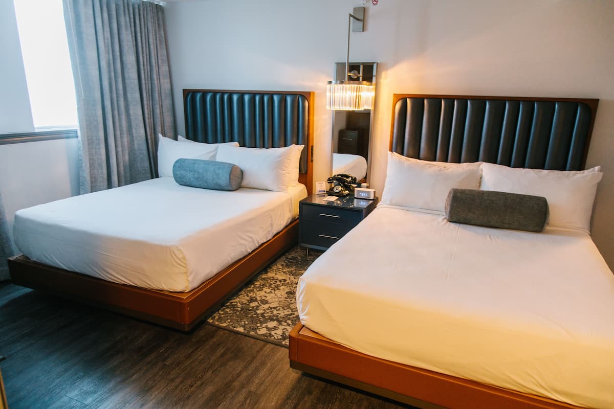 Quarto de hotel com duas camas de solteiro, uma mesa de cabeceira com abajur e uma pequena mesa com telefone.