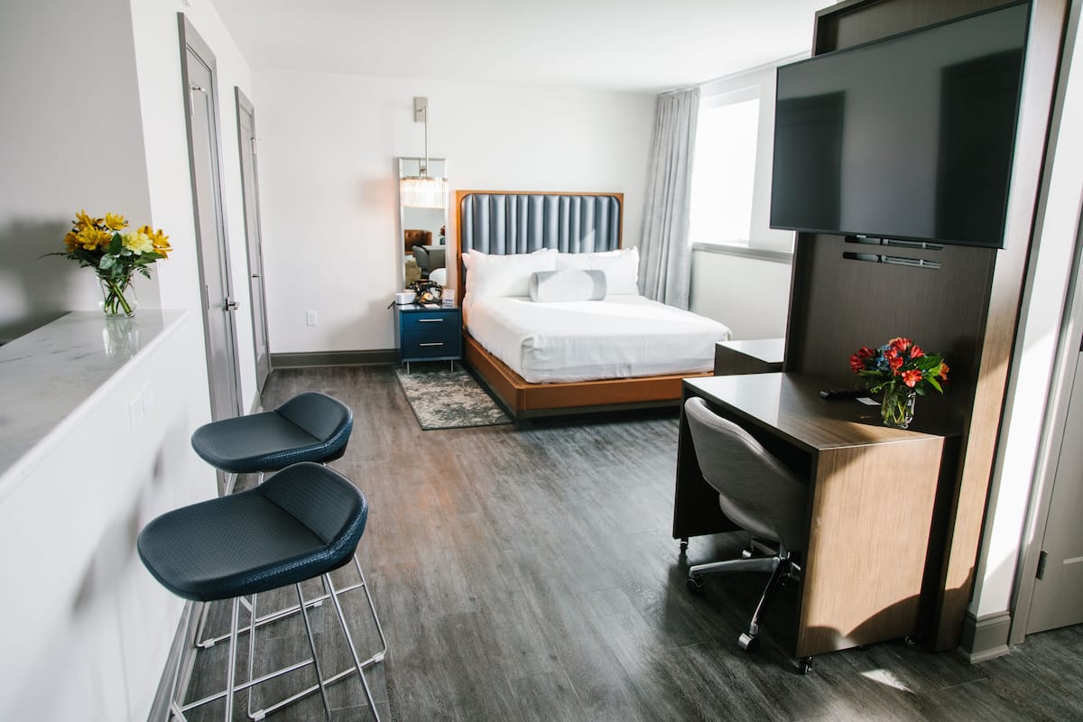 Quarto de hotel com cama, escrivaninha com cadeira, televisão e uma pequena mesa com um vaso de flores.