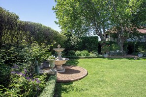 Um jardim bem cuidado com um gramado circular, uma fonte de pedra e diversas plantas e árvores.