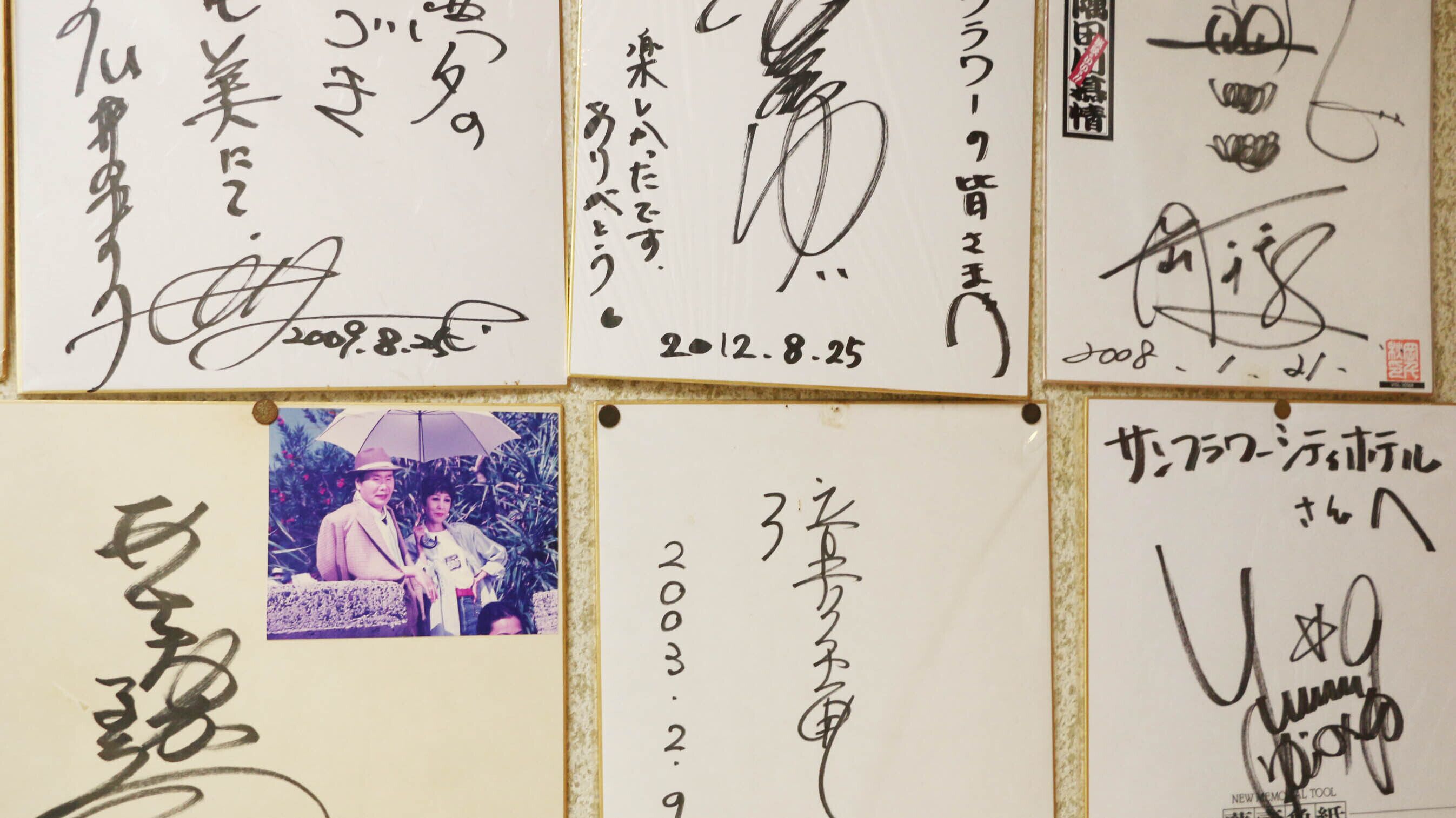 Notas manuscritas e assinaturas numa parede, com uma pequena fotografia de duas pessoas e um carimbo de data.