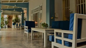 Um hall amplo com mobiliário branco e detalhes em azul, com ventilador de teto, janelas grandes e plantas em vasos.