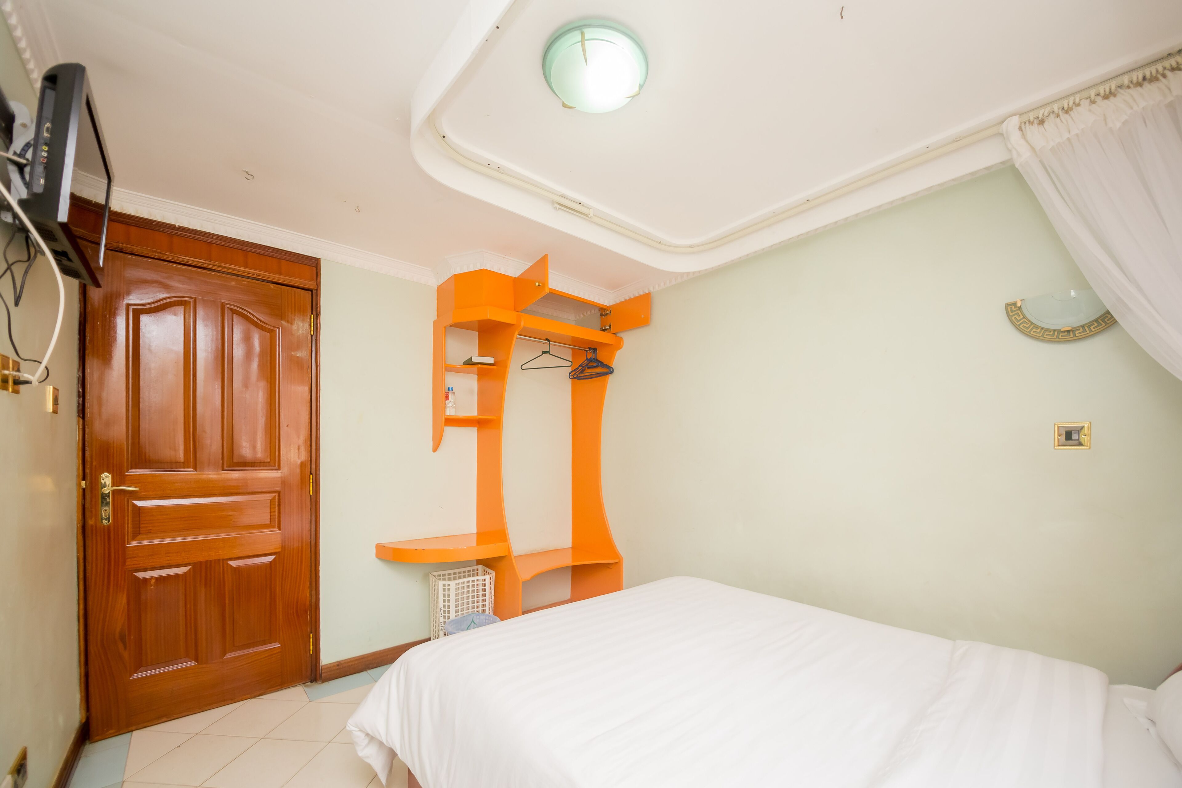 Quarto com porta de madeira, cama com roupa de cama branca, estante laranja fixada na parede e luminária no teto.