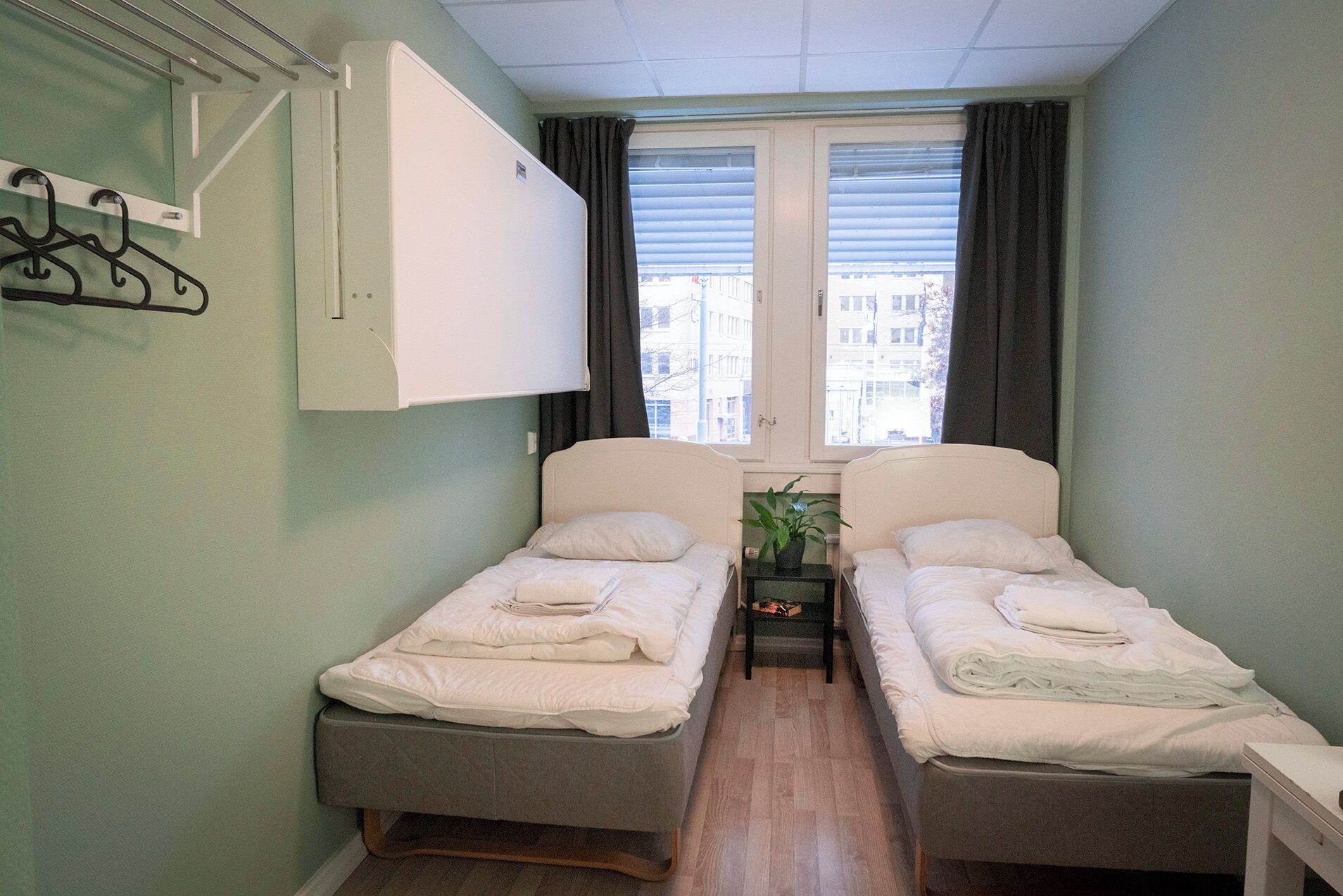 Quarto pequeno e estreito com duas camas de solteiro, uma mesinha com uma planta e uma janela com persianas.
