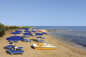 Uma praia com fileiras de guarda-sóis azuis e vermelhos, cadeiras de praia brancas e um barco amarelo.