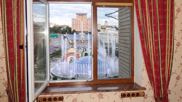 Uma janela com cortinas, vista de uma fonte e um edifício ao fundo.