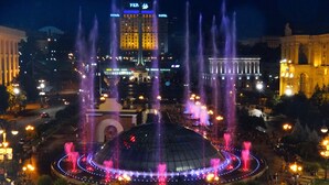 Uma grande fonte com jatos de água em padrão circular, cercada por uma estrutura em forma de cúpula. Ao fundo, um edifício com sinalização iluminada.