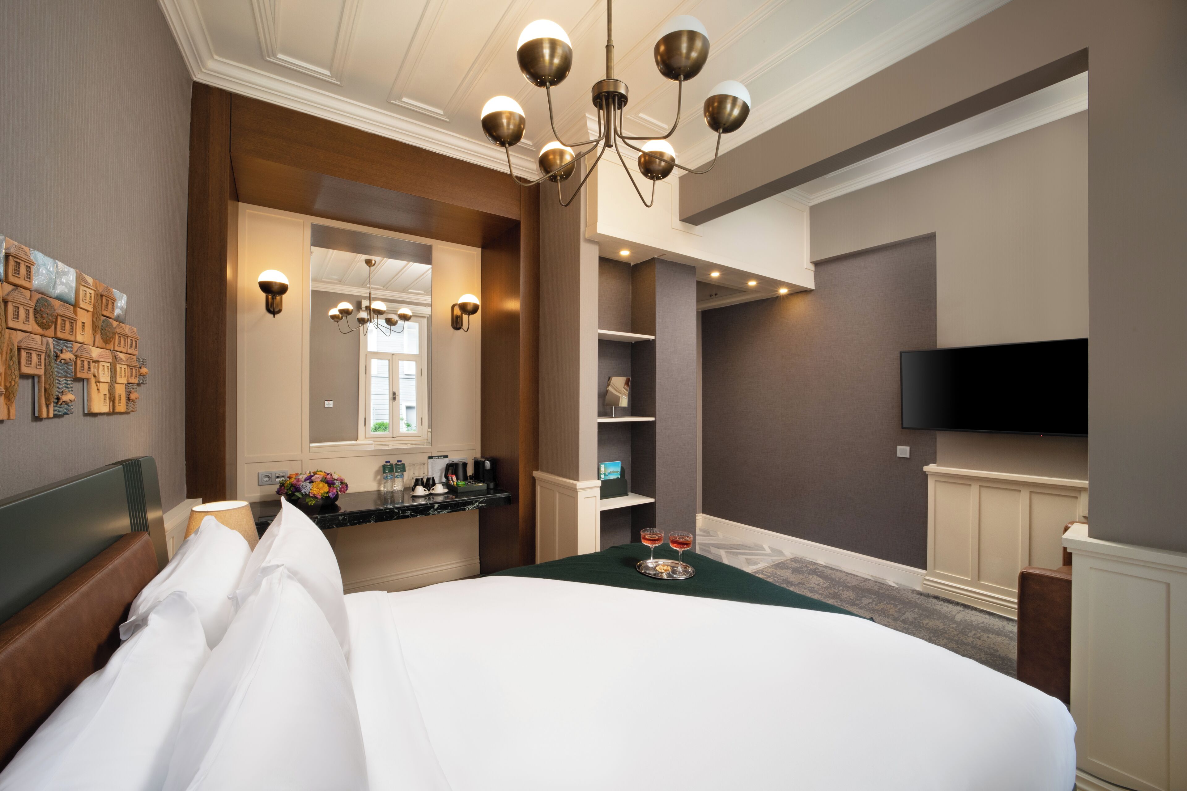 Quarto de hotel com uma cama grande, um candelabro, uma TV de tela plana, um espelho e uma mesa pequena com dois copos.