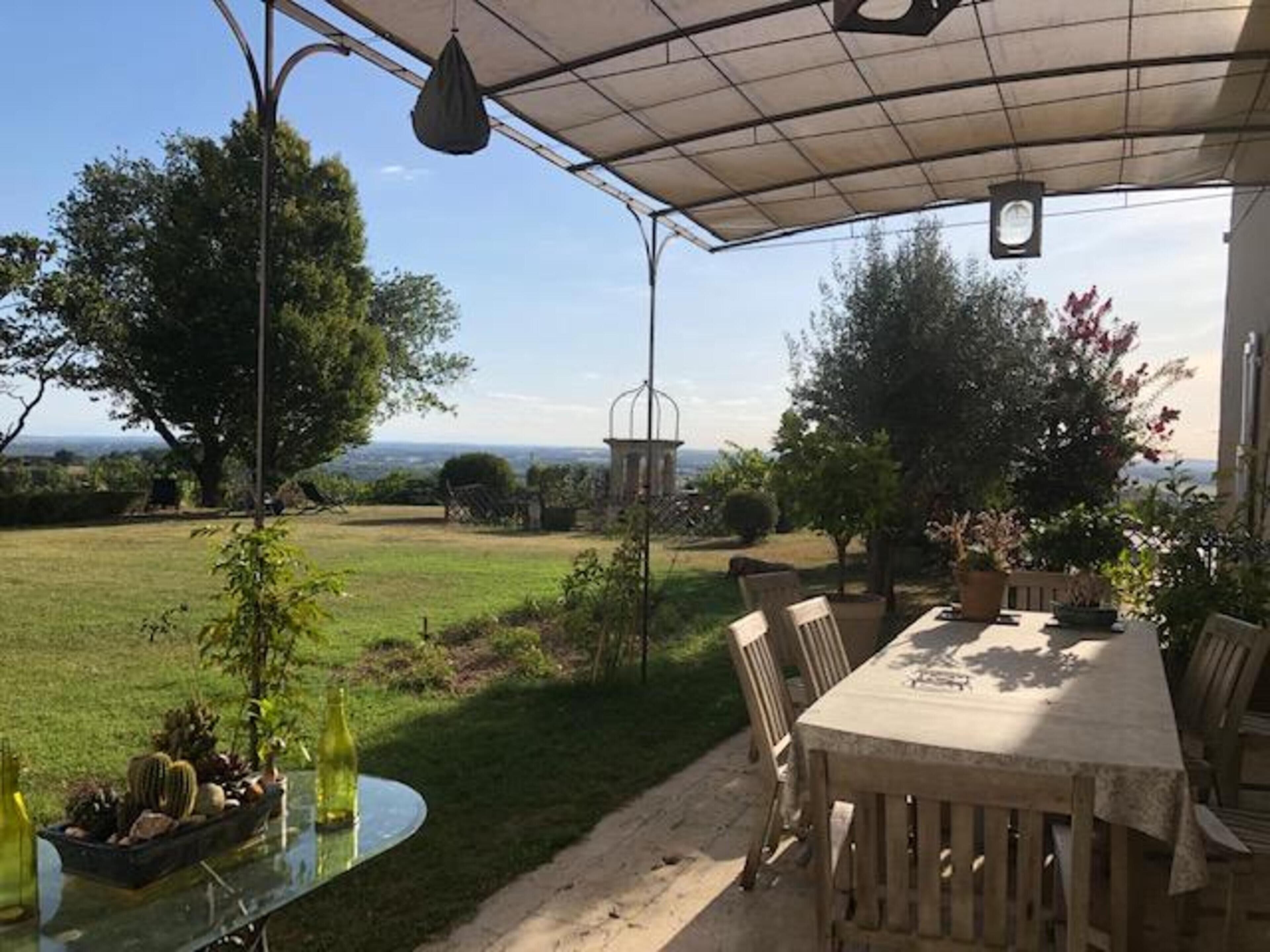 Um pátio externo com mesa e cadeiras, um candeeiro pendente e vista para o campo.
