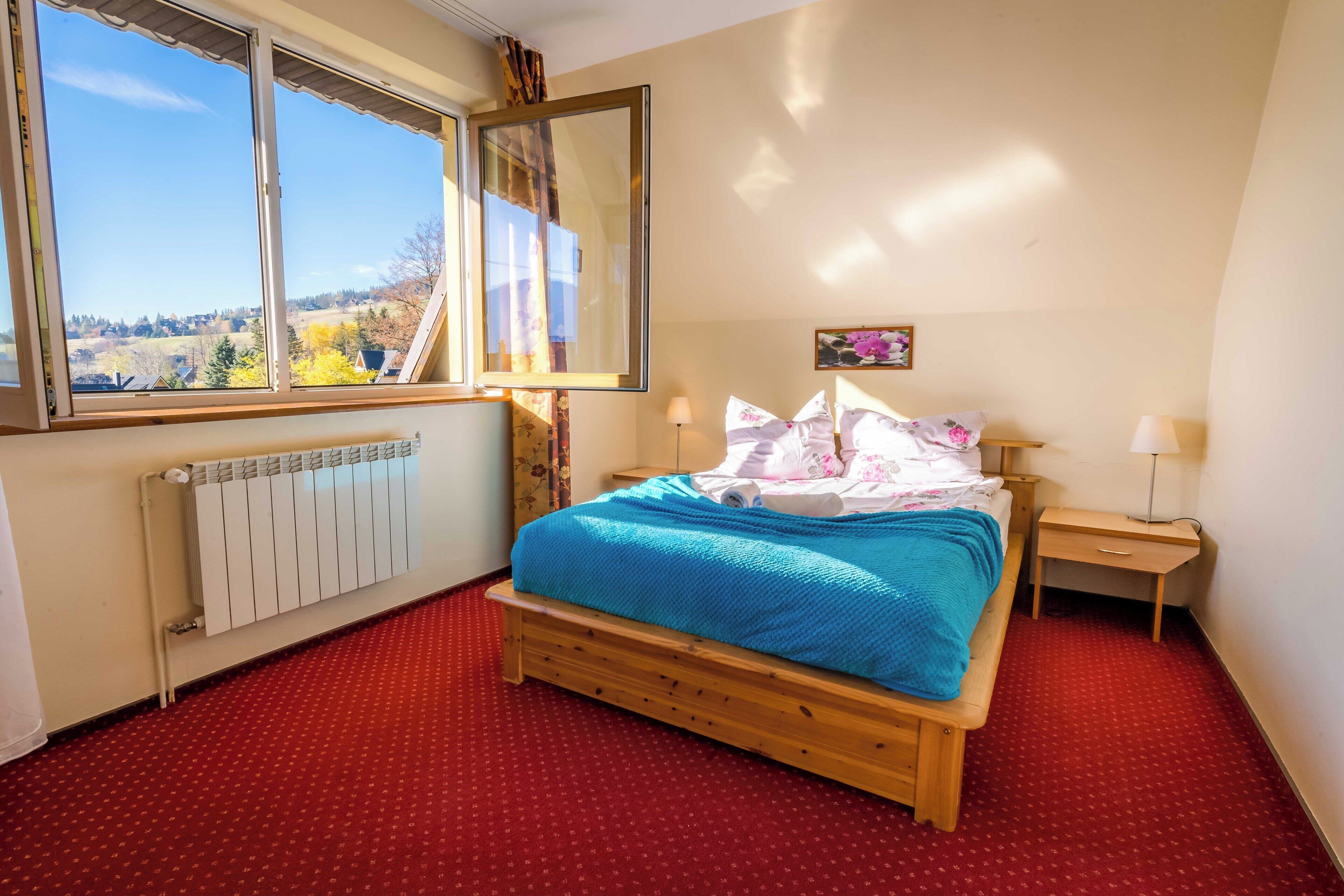 Quarto com cama de madeira, tapete vermelho, janela com vista, radiador e mesa de cabeceira.