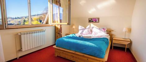 Quarto com cama de madeira, tapete vermelho, janela com vista, radiador e mesa de cabeceira.
