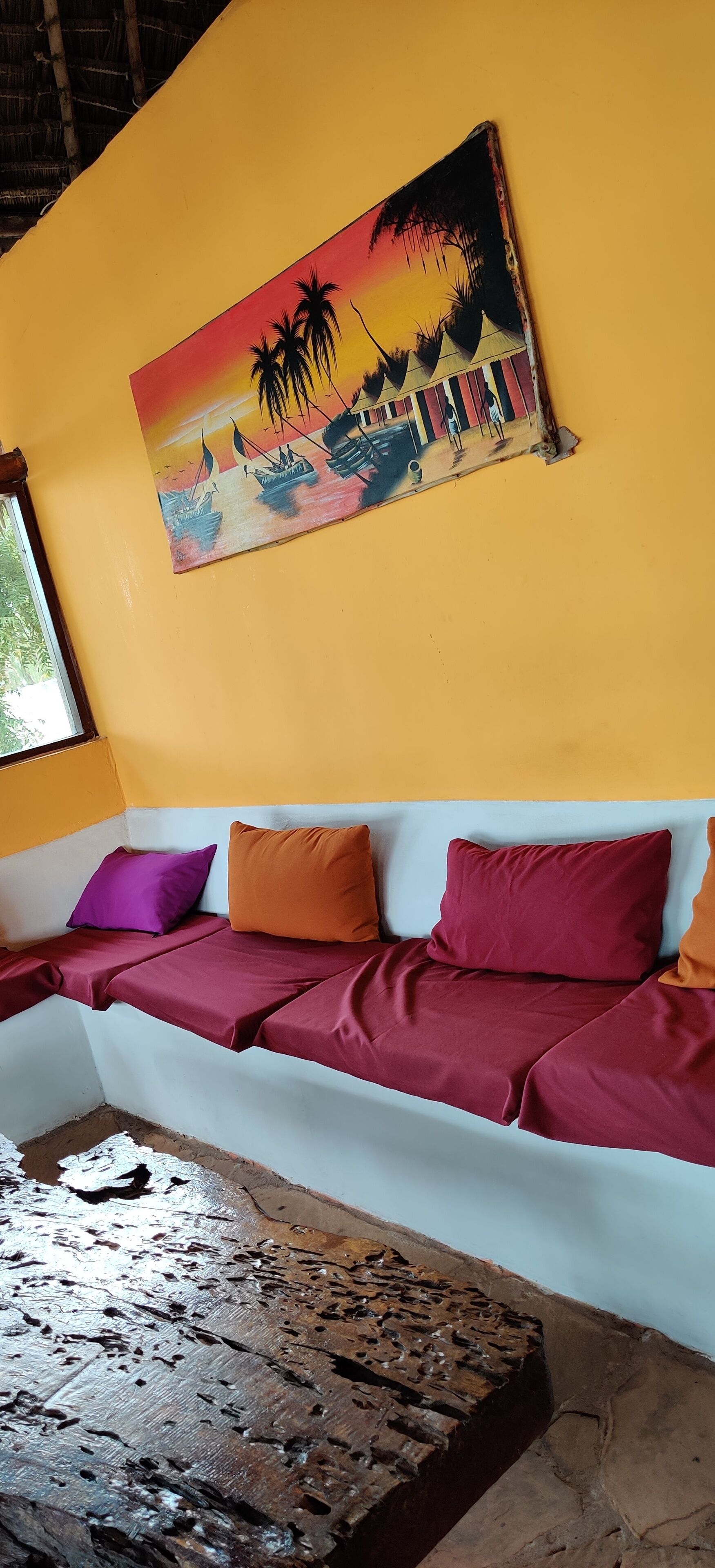 Um quarto com parede amarela, sofá branco com almofadas vermelhas e roxas, mesa de centro de madeira e uma pintura de uma cena de pôr do sol.