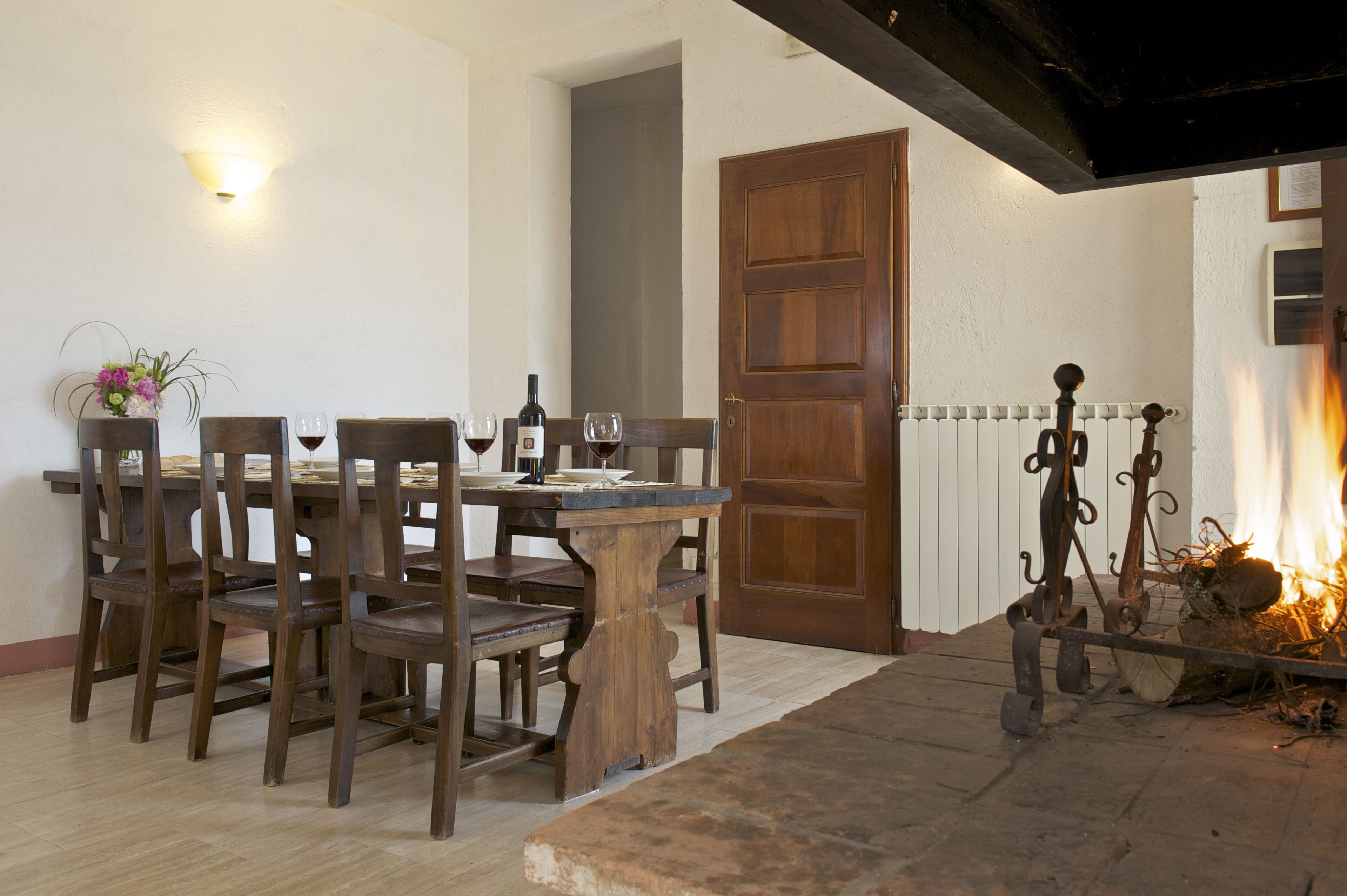 Uma sala de jantar com mesa e cadeiras de madeira, um vaso com flores e uma garrafa de vinho.