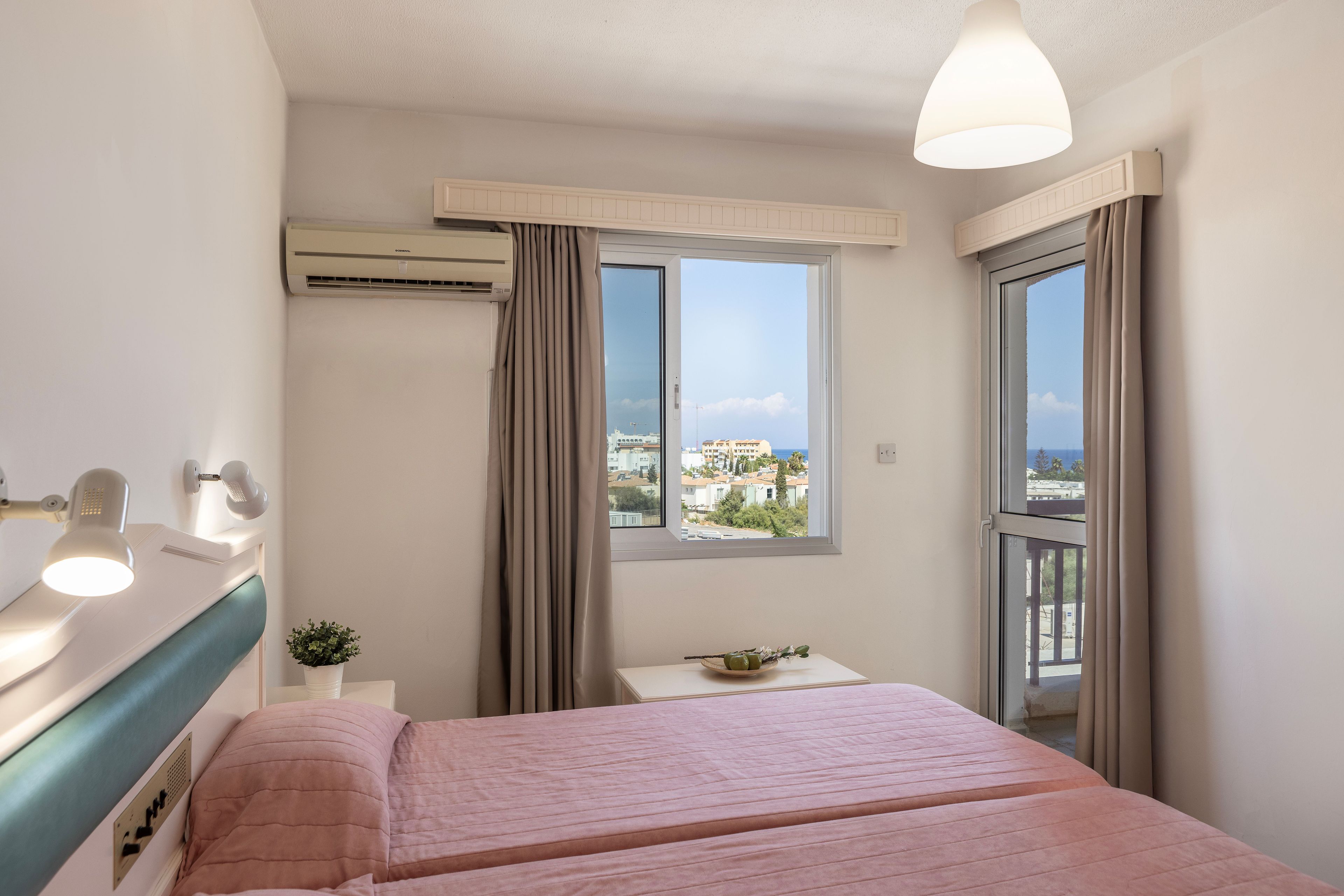 Quarto com cama, ar condicionado, abajur, pequena mesa com uma tigela e janela com cortinas.