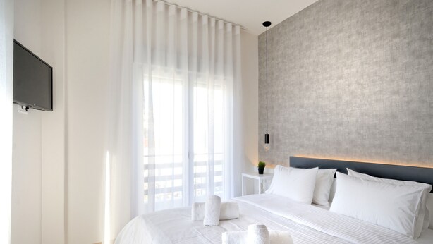 Quarto de hotel moderno com uma cama grande, roupa de cama branca, televisão de tela plana na parede e uma janela ampla com cortinas leves.