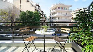 Uma varanda com uma mesa redonda e duas cadeiras de madeira, com vista para uma rua com carros estacionados e edifícios.