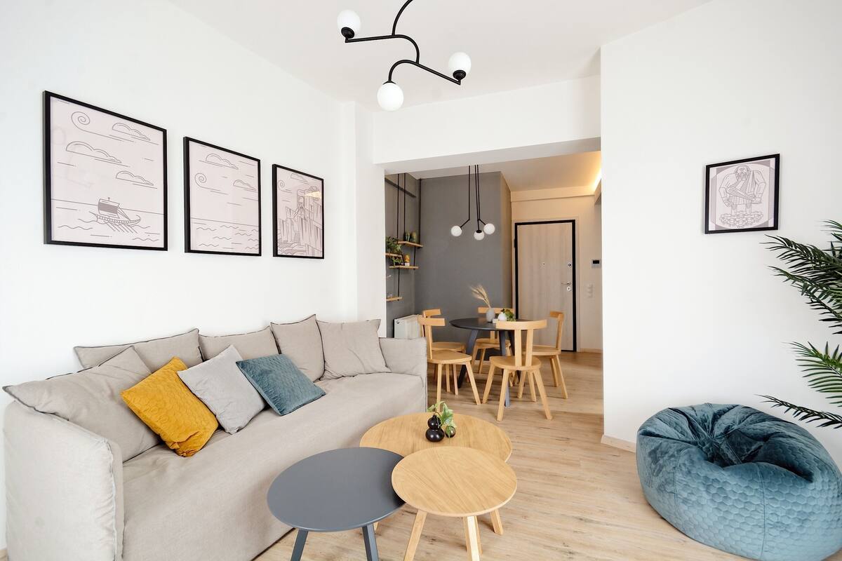 Uma sala de estar moderna com um sofá, mesa de centro e um puff. Há quadros nas paredes e uma área de jantar ao fundo.