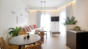 Uma sala de estar moderna com uma mesa de jantar redonda, cadeiras de vime, um sofá com almofadas, uma TV na parede e uma planta em vaso.