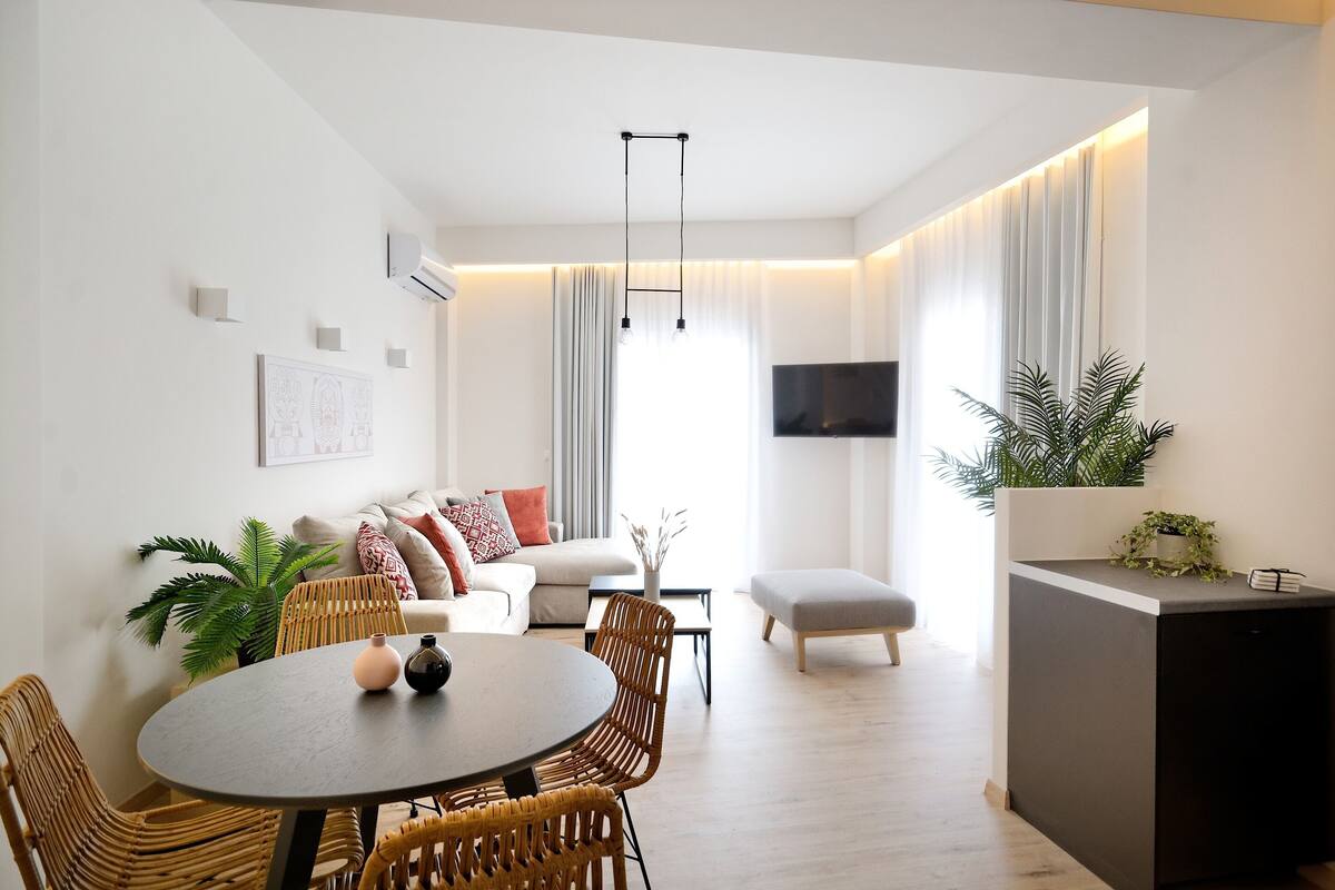 Uma sala de estar moderna com uma mesa de jantar redonda, cadeiras de vime, um sofá com almofadas, uma TV na parede e uma planta em vaso.