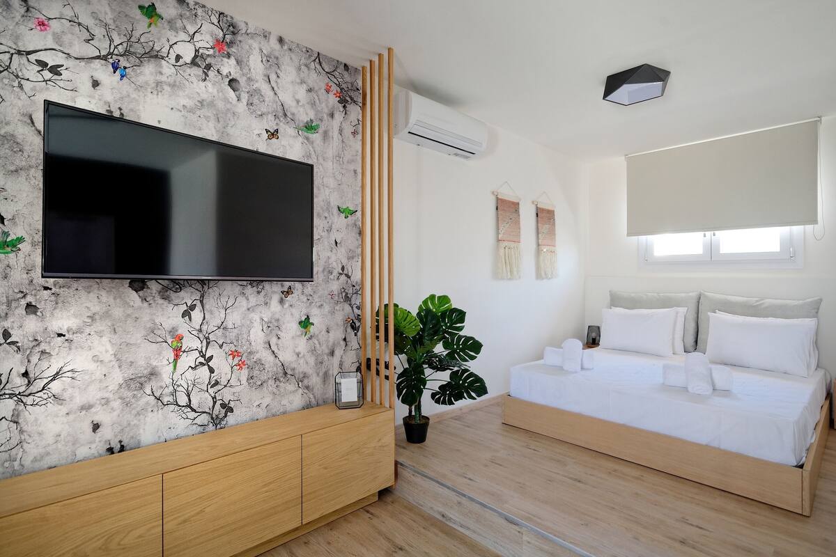 Quarto moderno com televisão de tela plana grande, um vaso de planta, cama com roupa de cama branca e janela com persiana.